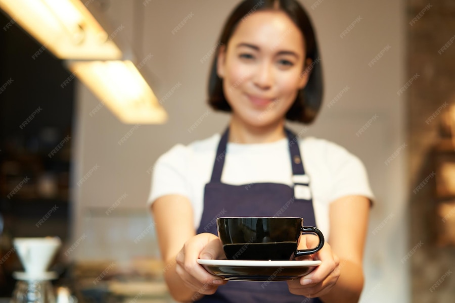 Menina asiática sorridente, barista de uniforme de avental, dando-lhe ... Menina asiática sorridente, barista de uniforme de avental, dando-lhe ...
