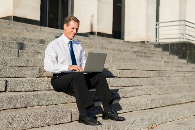 Free Photo | Smiling mature businessman sitting on staircase using laptop Free Photo | Smiling mature businessman sitting on staircase using laptop