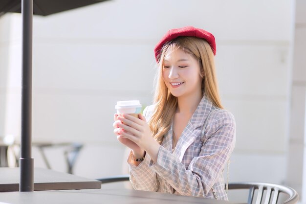 スーツとブロンズの髪の赤い帽子をかぶったアジア人女性が、コーヒーカップを持ちながら店の椅子に座る プレミアム写真